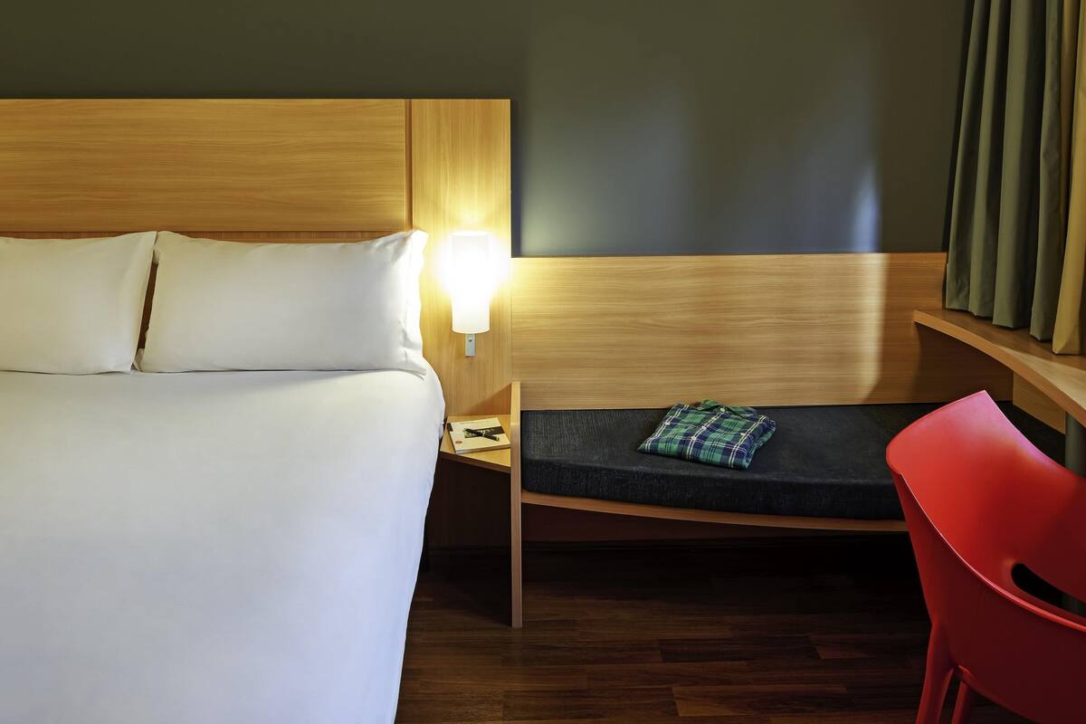 Quarto de hotel com cama, mesa de cabeceira com luminária, pequeno banco com almofada e uma cadeira vermelha.