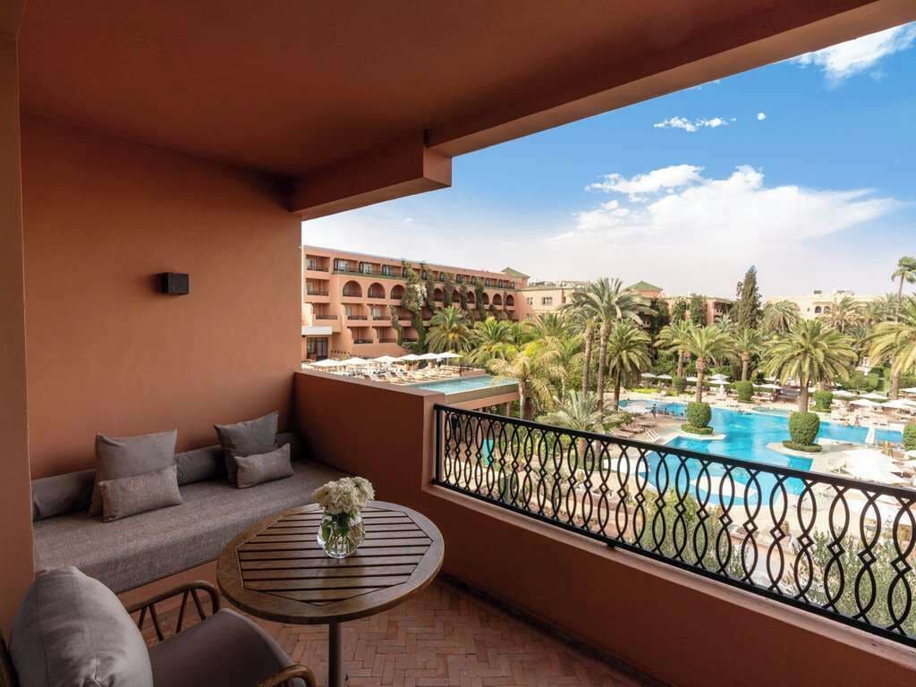Varanda com sofá, mesa redonda com um vaso e vista para uma piscina e edifícios de hotel.
