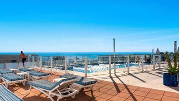 Uma área de piscina no terraço com espreguiçadeiras e guarda-corpo de vidro com vista para o mar.