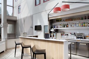 Uma área de bar moderna com balcão de madeira, dois taburetes pretos, uma televisão na parede e diversos copos e garrafas em prateleiras.