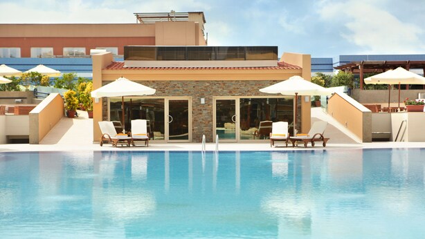 Piscina com espreguiçadeiras e guarda-sóis, rodeada por um edifício com telhado em azulejos e paredes de pedra.