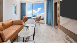 Quarto de hotel moderno com sofá de couro, uma mesinha redonda com um vaso de flores, televisão de tela plana e uma janela grande com vista para o céu.