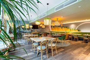 Um restaurante com mesas e cadeiras de madeira, luminárias pendentes e uma área de estar acolchoada em tons de verde.
