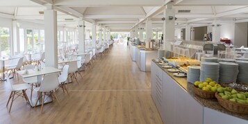 Uma ampla área de buffet com mesas e cadeiras brancas, um balcão extenso com comida e bebidas, e grandes janelas que permitem entrada de luz natural.
