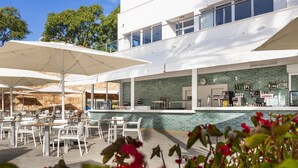 Uma área de estar ao ar livre com mesas brancas e guarda-sóis, um bar com painel de azulejos verdes e uma parede de pedra ao fundo.