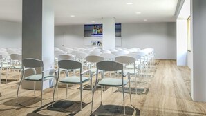 Uma sala de conferências com fileiras de cadeiras brancas, piso de madeira e um pilar grande.