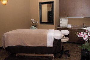 Uma mesa de massagem com toalha branca, uma cadeira pequena, uma pia e um espelho num quarto com paredes bege e um armário de madeira.