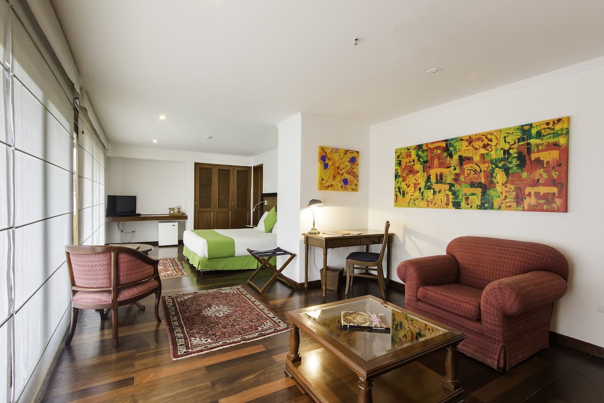 Uma sala de estar espaçosa com uma janela grande, um sofá verde, uma poltrona vermelha, uma mesa de centro de madeira e uma escrivaninha com uma luminária.