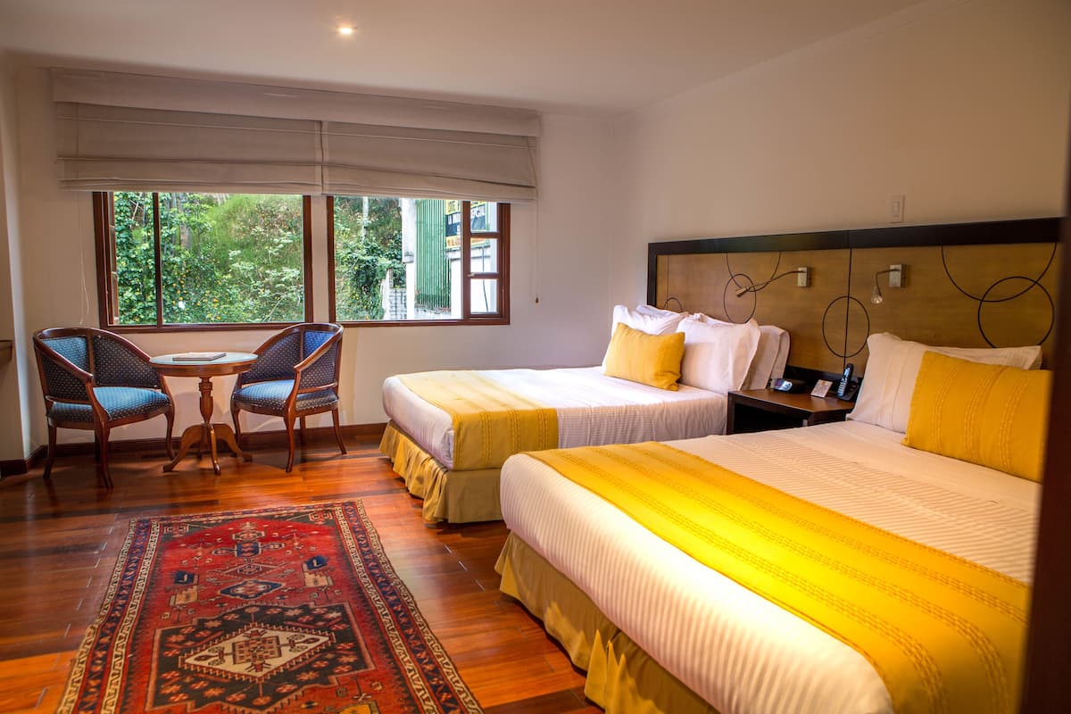 Quarto de hotel com duas camas, uma mesinha com cadeiras e vista para o verde lá fora.