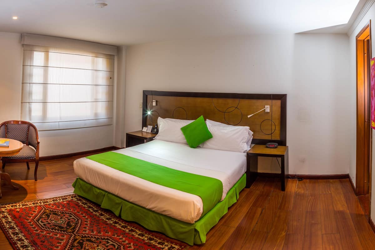 Quarto de hotel com uma cama grande, uma cadeira, uma mesinha, uma janela com persianas e um quadro na parede.