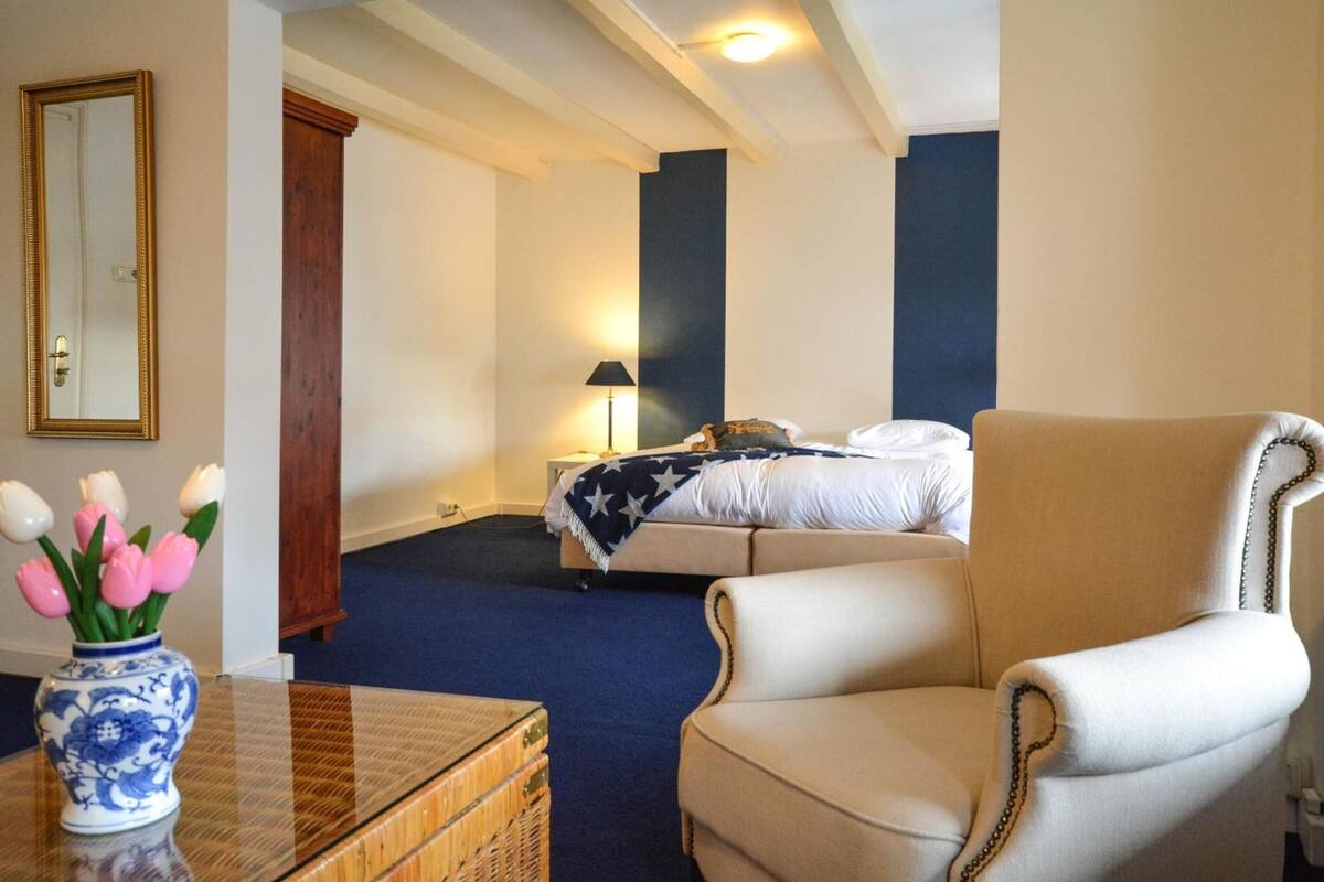 Quarto de hotel com uma cama, uma cadeira, penteadeira de madeira, um vaso com flores e um espelho na parede.