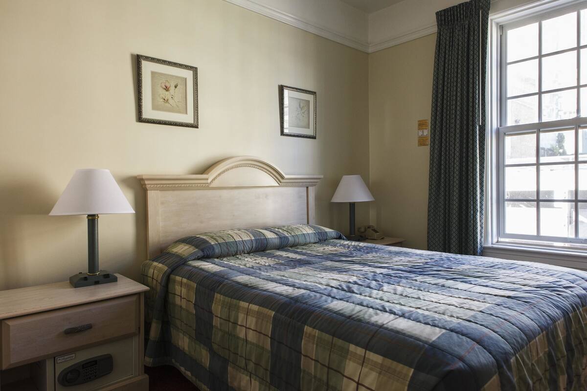 Quarto de hotel com uma cama, duas mesas de cabeceira, luminárias, uma cabeceira e duas fotos emolduradas na parede.