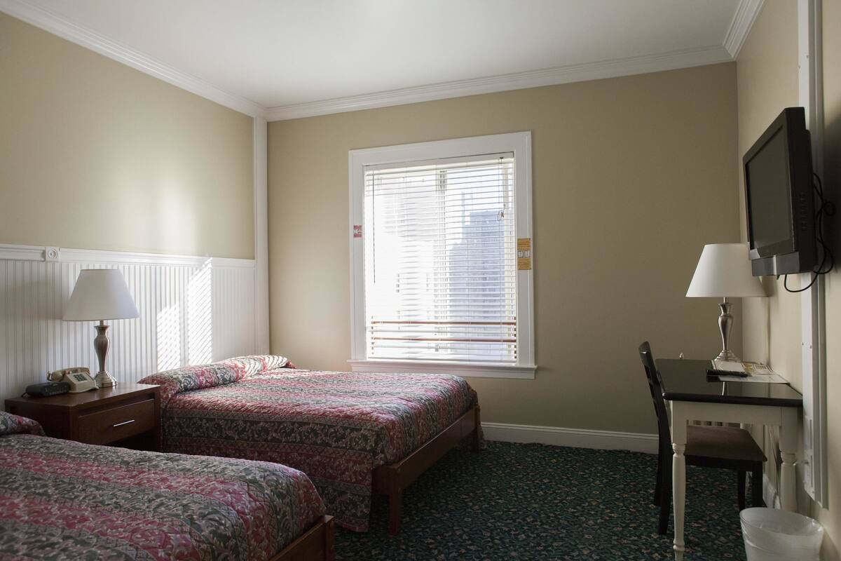 Quarto de hotel com duas camas, uma escrivaninha, uma cadeira, uma televisão e uma janela com persianas.