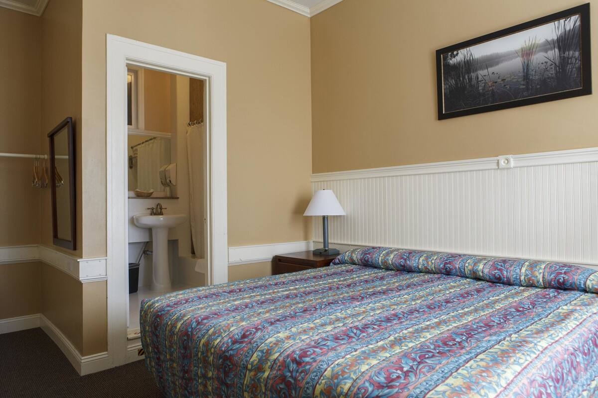 Quarto de hotel com cama, mesa de cabeceira, luminária, quadro na parede e banheiro visível por uma porta aberta.