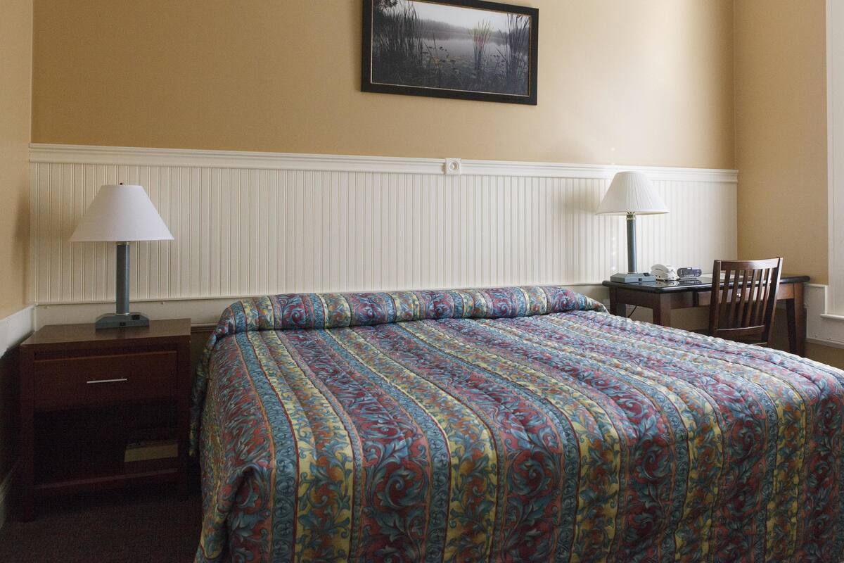 Quarto de hotel com uma cama, duas mesas de cabeceira com luminárias, uma cadeira e um quadro na parede.