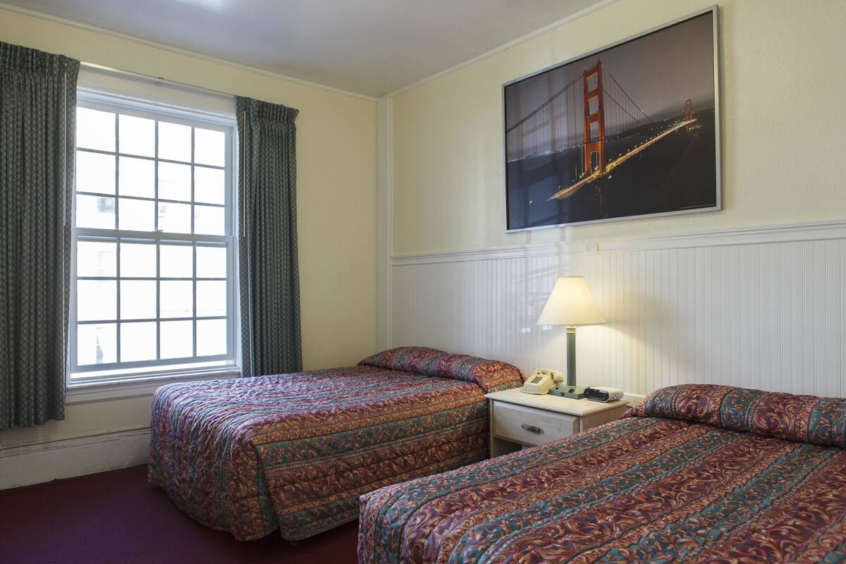 Quarto de hotel com duas camas, janela com cortinas, mesa de cabeceira com abajur e um quadro da Ponte Golden Gate.