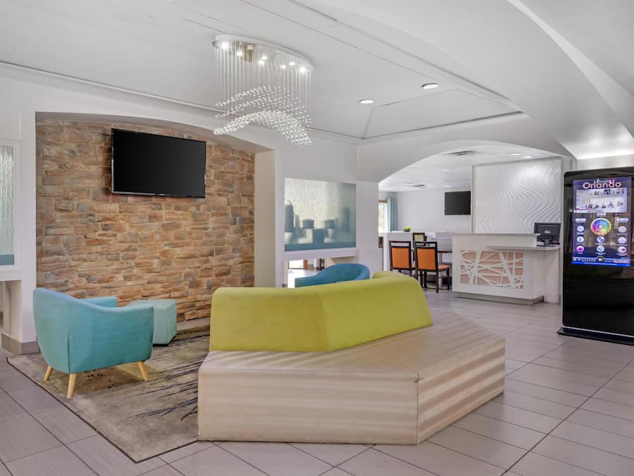 Um saguão de hotel moderno com parede de pedra, televisão de tela plana, sofá amarelo, poltronas verde-água, balcão de recepção e um quiosque de informação digital.