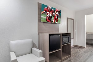 Quarto de hotel com televisão de tela plana na parede, uma pequena geladeira preta e uma poltrona cinza.