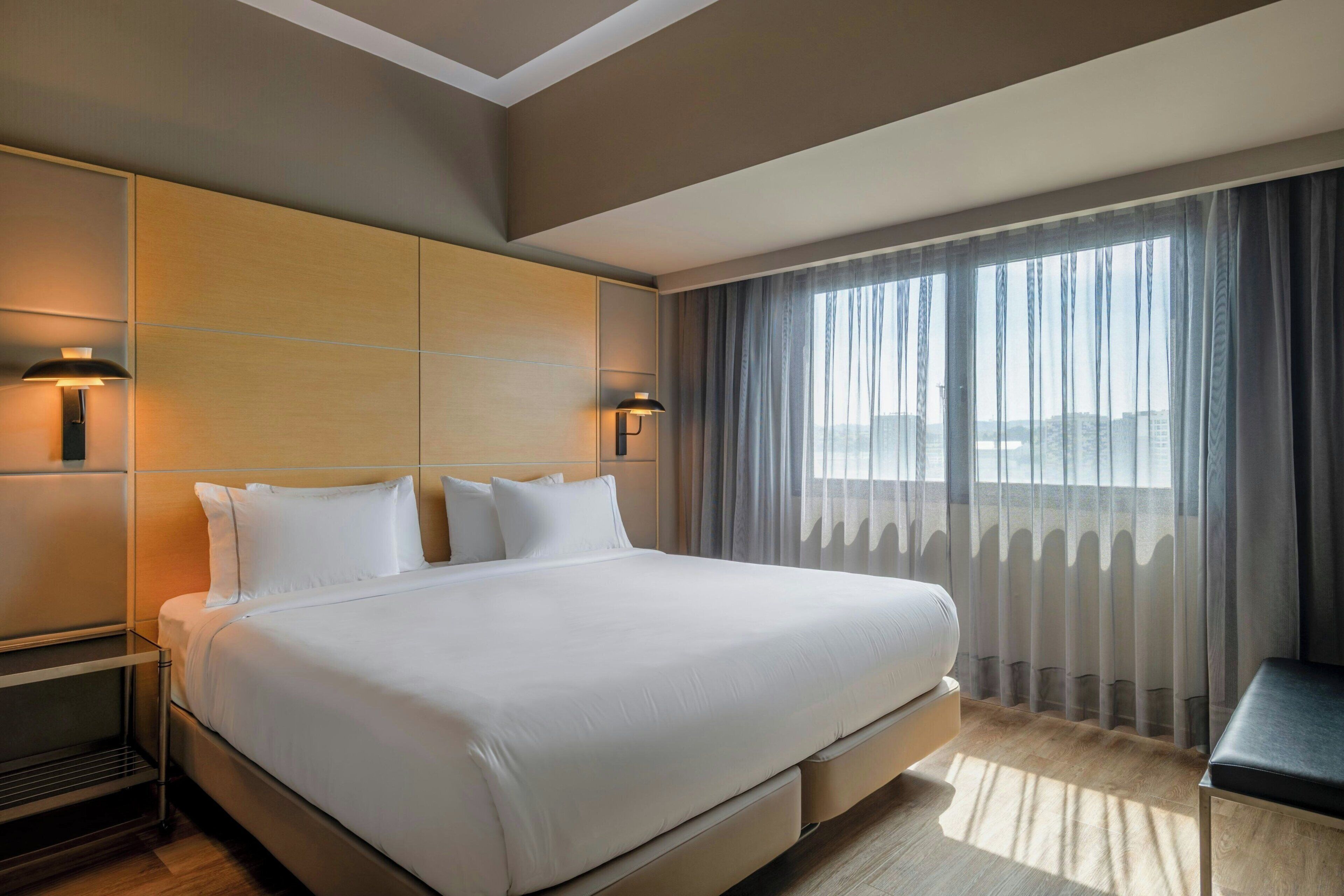 Quarto de hotel com uma cama grande, abajures, vista da cidade através de cortinas finas e piso de madeira.