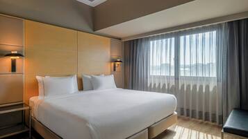 Quarto de hotel com uma cama grande, abajures, vista da cidade através de cortinas finas e piso de madeira.