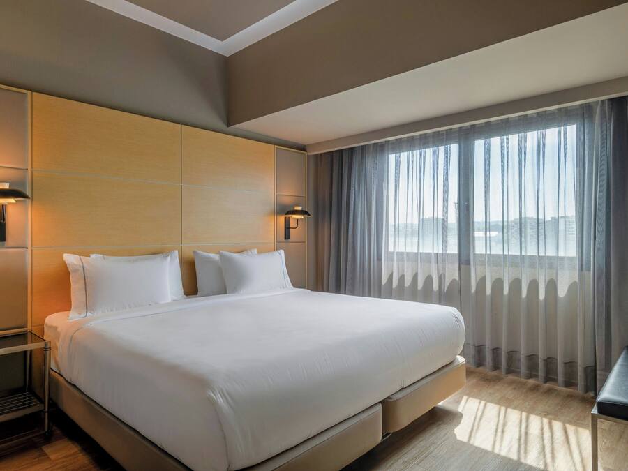 Quarto de hotel com uma cama grande, abajures, vista da cidade através de cortinas finas e piso de madeira.