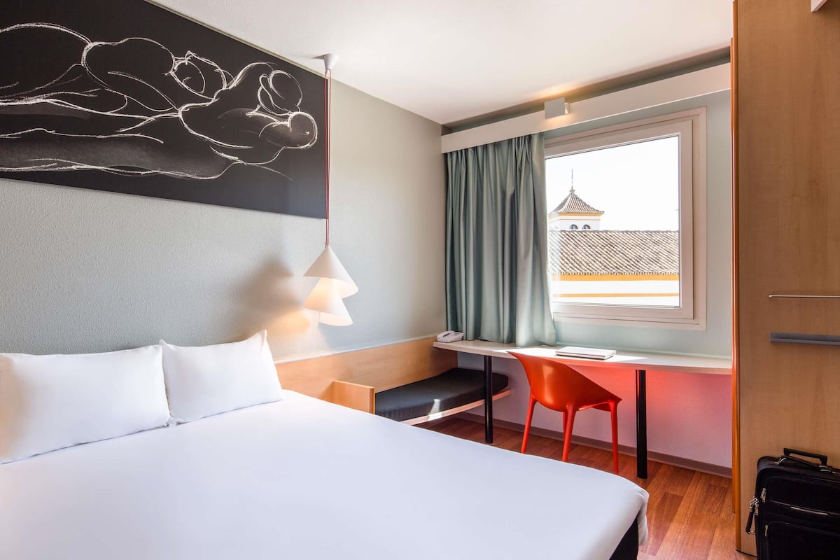 Quarto de hotel com uma cama grande, uma escrivaninha com cadeira, uma janela com cortinas e uma luminária na parede.