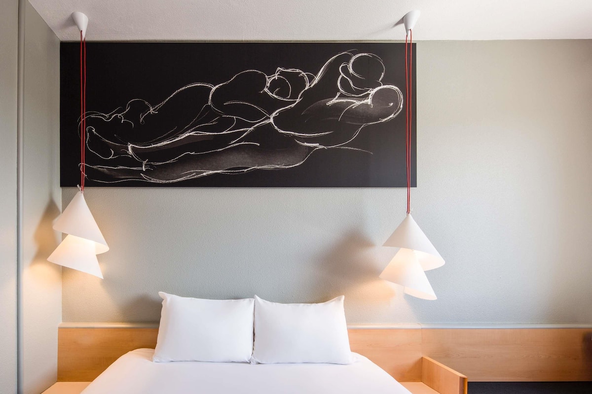 Quarto de hotel com cama, duas luminárias de cabeceira, um painel de cabeceira de madeira e uma obra de arte em preto e branco na parede.