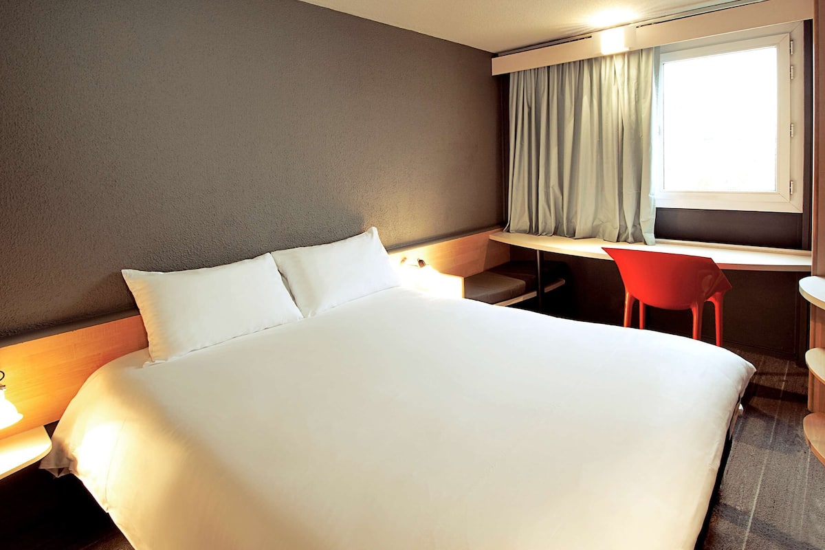Quarto de hotel com cama, escrivaninha, cadeira e janela com cortinas.