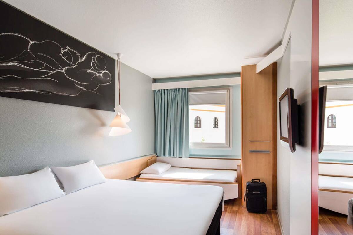 Quarto de hotel com duas camas, uma janela grande com cortinas, uma TV fixada na parede e uma luminária no teto.