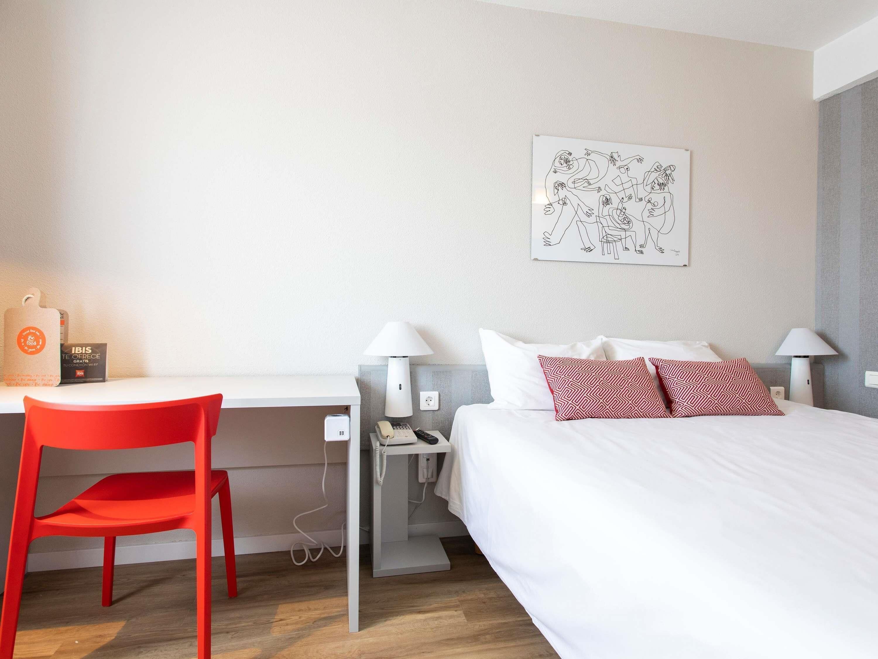 Quarto de hotel com cama, escrivaninha, cadeira, abajur, telefone e uma obra de arte na parede.