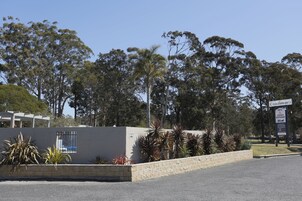 Um hotel com uma placa, uma área pavimentada e uma fileira de árvores ao fundo.