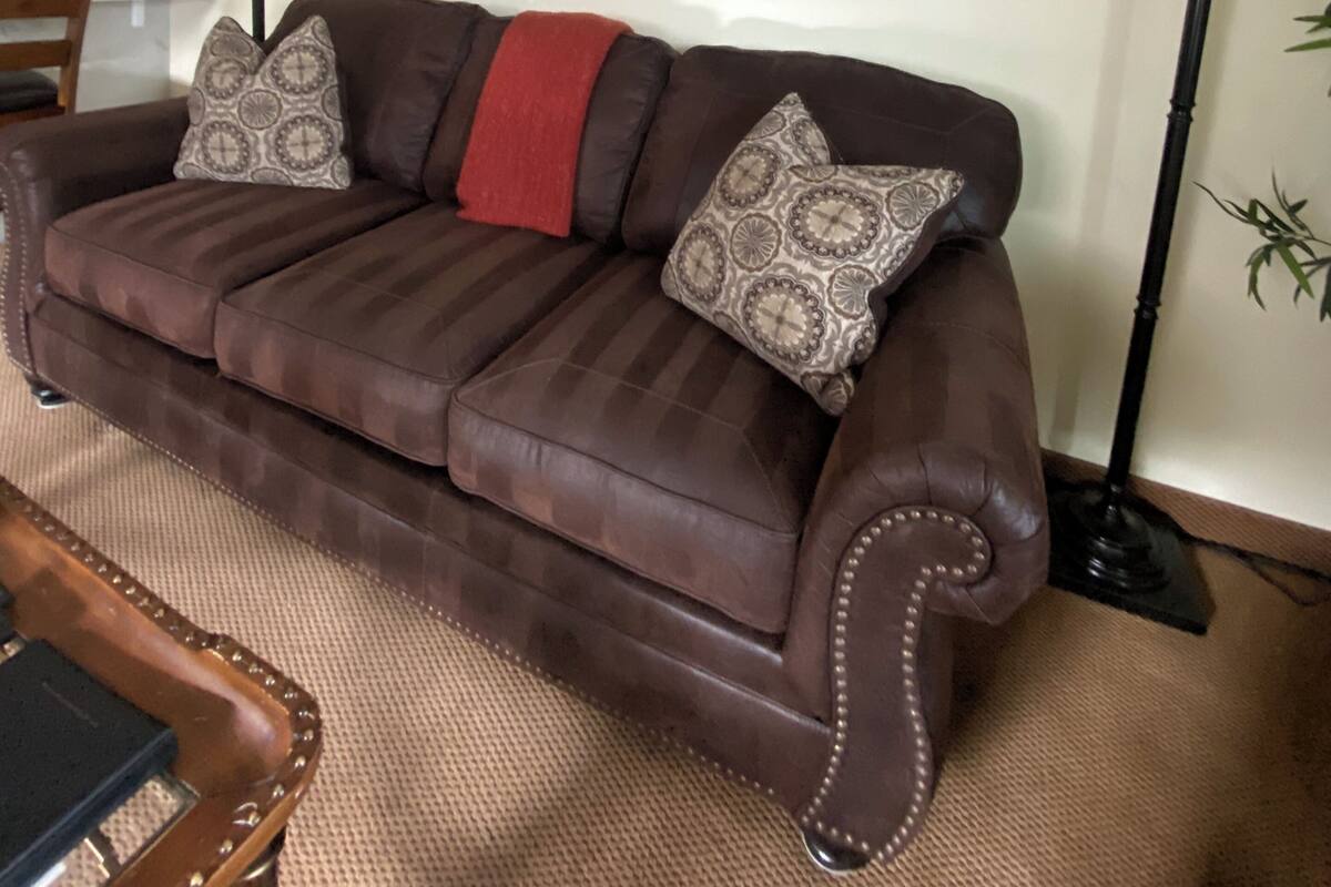 Um sofá de couro marrom com almofadas estampadas, um cobertor vermelho e um quadro de montanhas na parede.