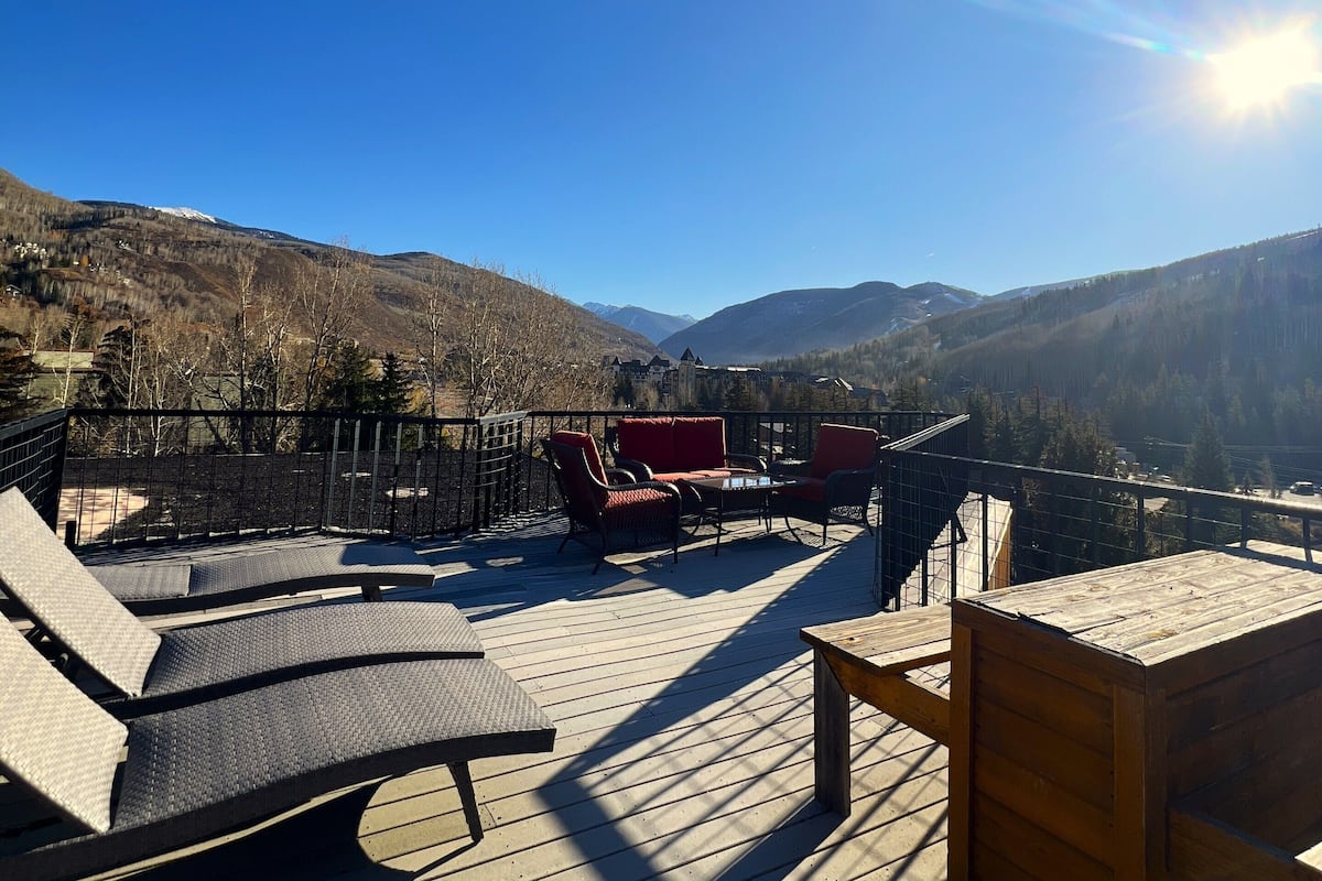 Um deck de madeira com cadeiras de descanso e uma mesa, com vista para uma paisagem montanhosa.
