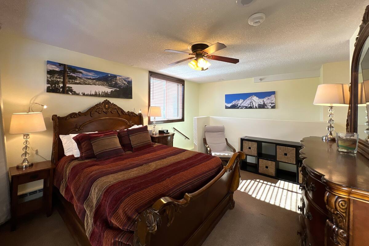 Quarto com cama de madeira, duas mesas de cabeceira com luminárias, um ventilador de teto e duas pinturas de montanhas penduradas na parede.
