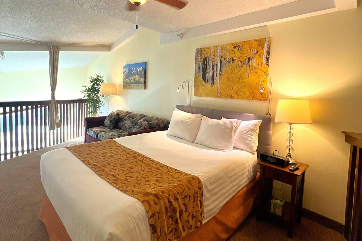 Quarto de hotel com uma cama grande, um ventilador de teto, um quadro na parede e uma mesa de cabeceira com abajur e controle remoto.