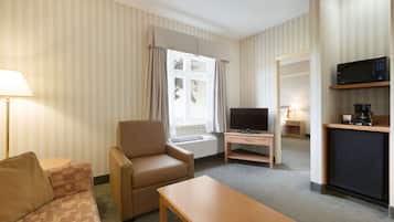 Quarto de hotel com sofá, poltrona, televisão, mesa pequena e cozinha compacta.