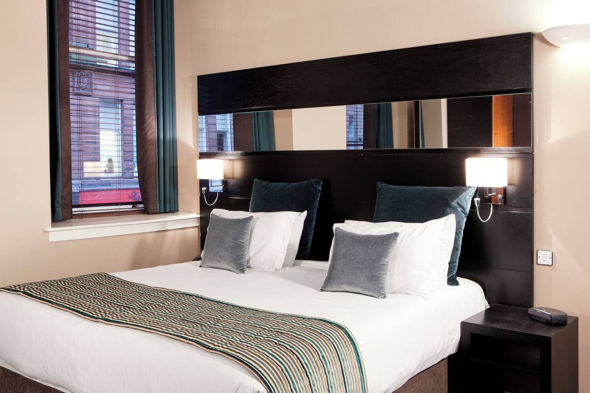 Quarto de hotel com uma cama grande, duas mesas de cabeceira, cabeceira com espelho e janela com persianas.