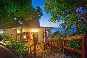 Uma casa de madeira com uma escadaria de pedra que leva a um alpendre, rodeada por vegetação exuberante e vista para o oceano.