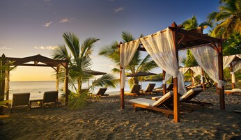 Uma praia com cabanas de madeira, palmeiras e espreguiçadeiras.