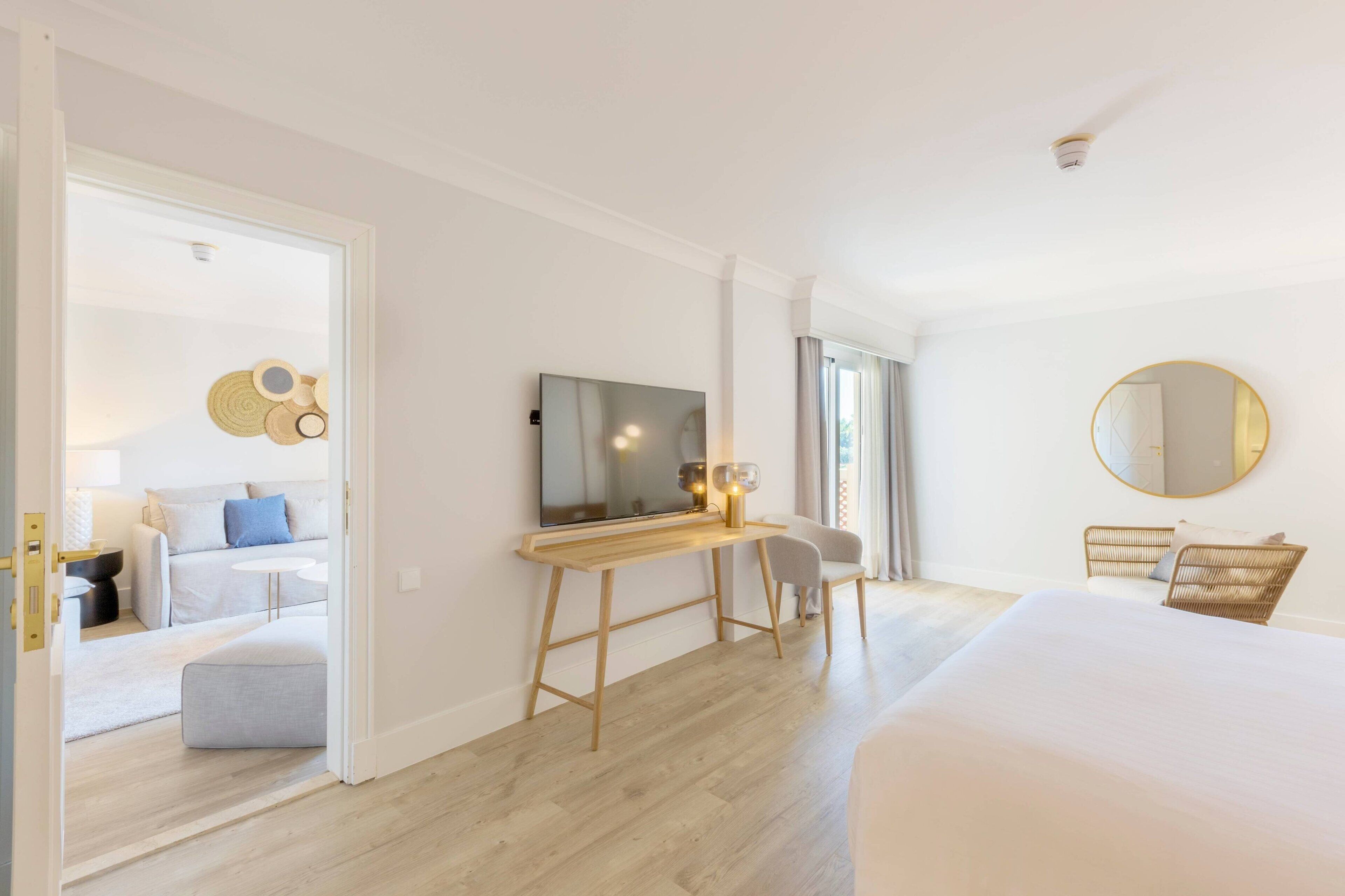 Quarto de hotel com cama, escrivaninha com televisão, cadeira, espelho redondo e vista para outro quarto.