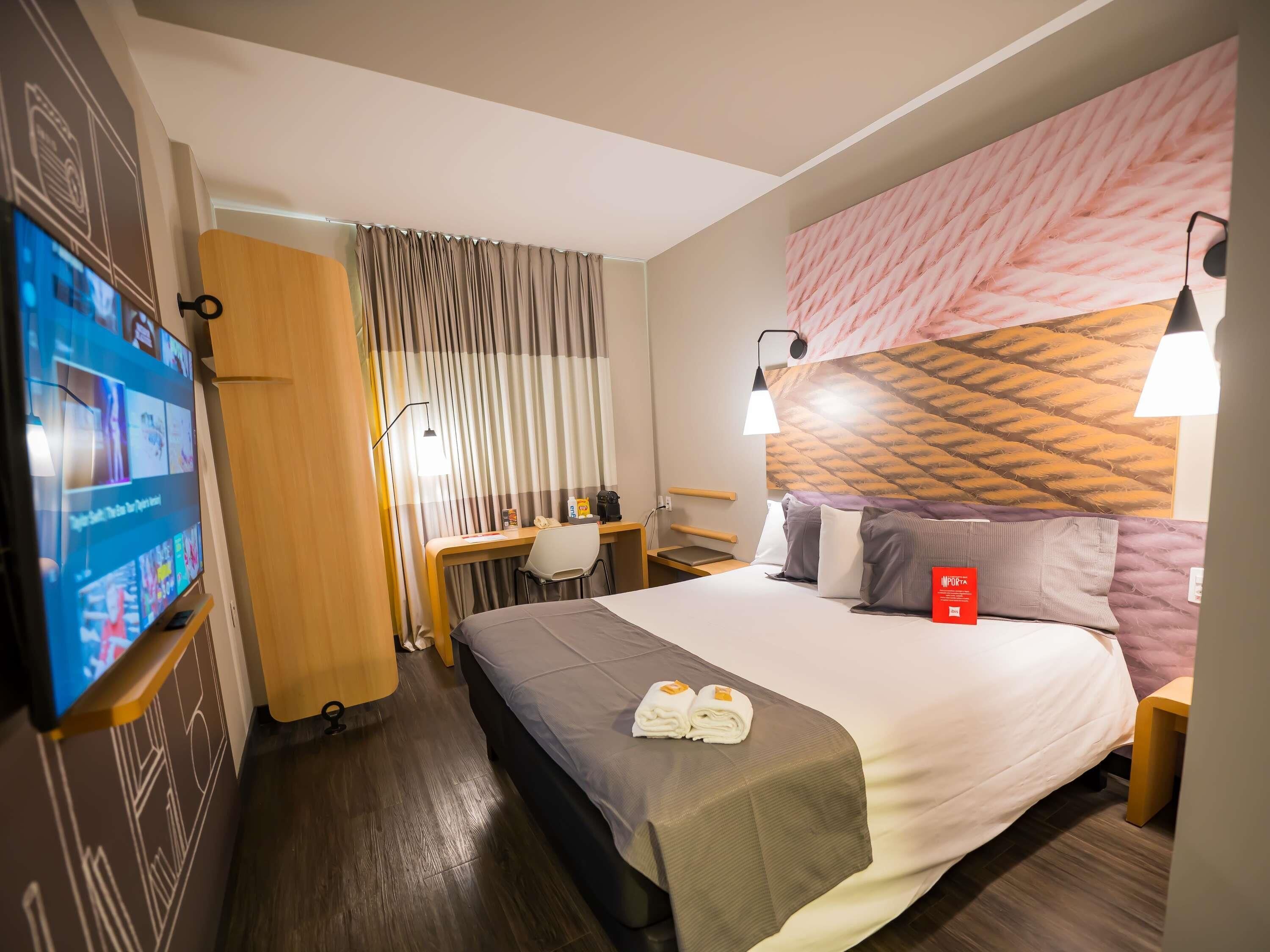 Quarto de hotel com cama, escrivaninha, cadeira, televisão e luminária na parede.