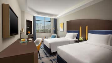 Quarto de hotel com duas camas, uma escrivaninha, uma cadeira, uma televisão e janela com vista para a cidade.