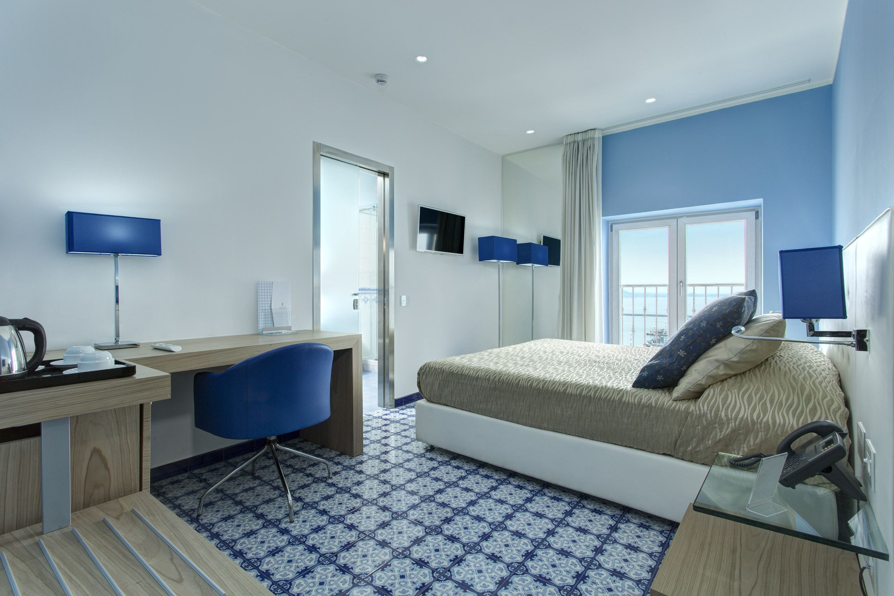 Quarto de hotel moderno com cama, escrivaninha e cadeira, uma janela grande e vista para a cidade.