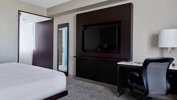 Quarto de hotel com uma cama, uma escrivaninha, uma cadeira, uma televisão e um abajur.