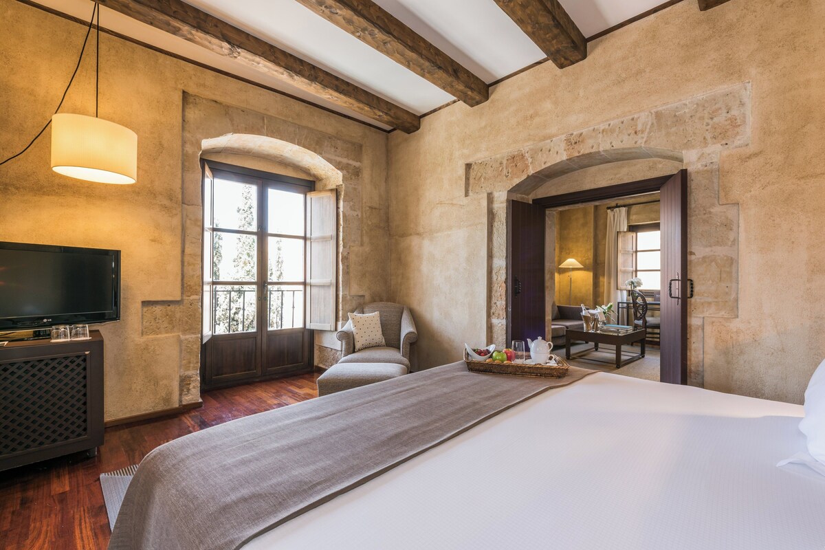 Quarto com uma cama grande, televisão, cadeira, mesa com uma bandeja de objetos e vista por uma porta arqueada.