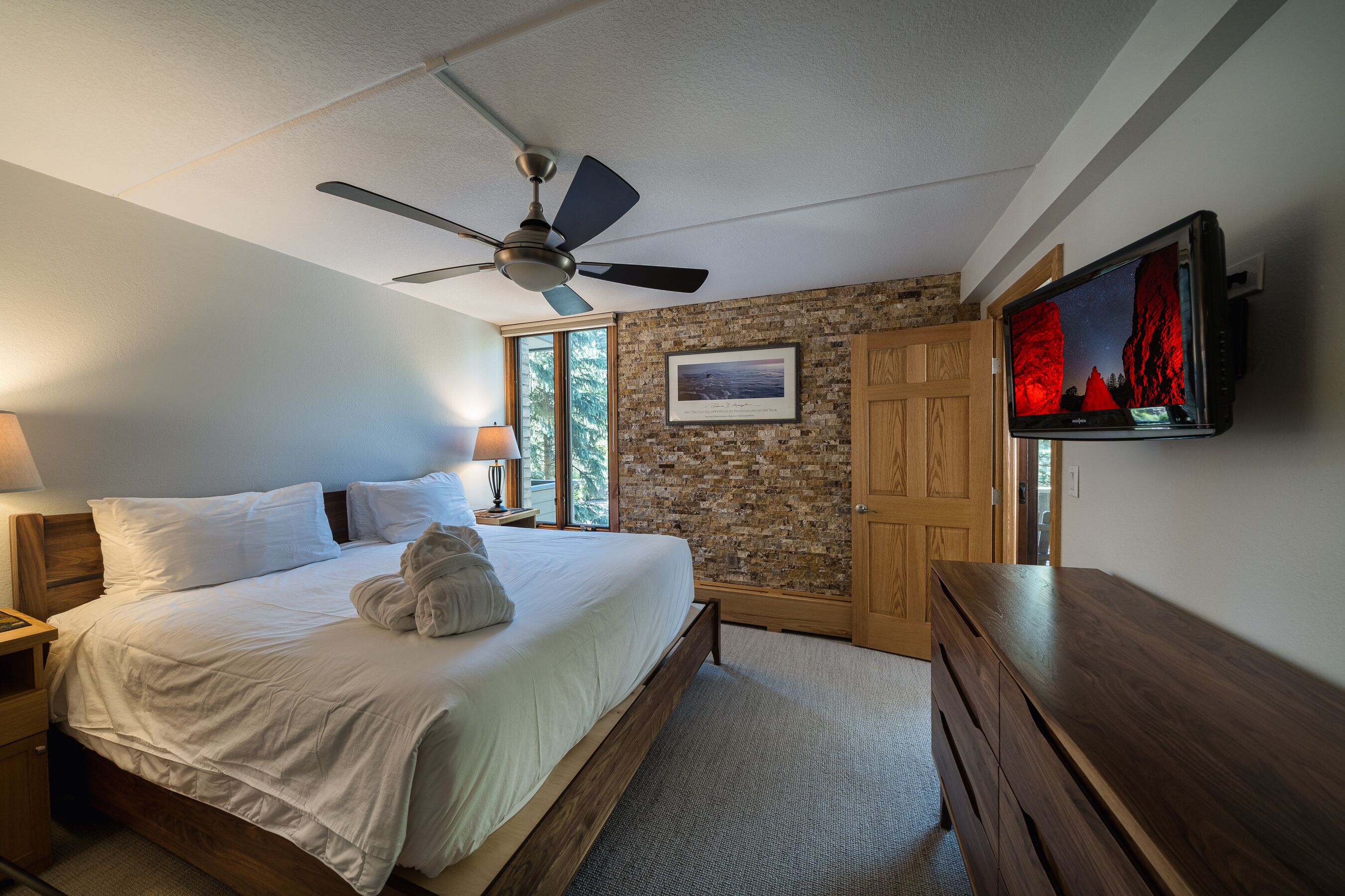 Quarto com cama, ventilador de teto, televisão, cômoda de madeira e parede de tijolos.