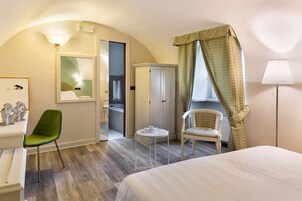Quarto de hotel com cama, escrivaninha com cadeira, mesinha, guarda-roupa e banheiro visível por uma porta arqueada.