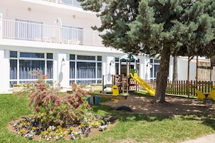 Espaço infantil com escorrega e estrutura de escalada, rodeado por um jardim com flores coloridas e uma grande árvore.