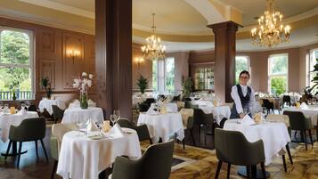 Uma sala de jantar formal com várias mesas preparadas para o serviço, um garçom de uniforme e grandes janelas com vista para o exterior.
