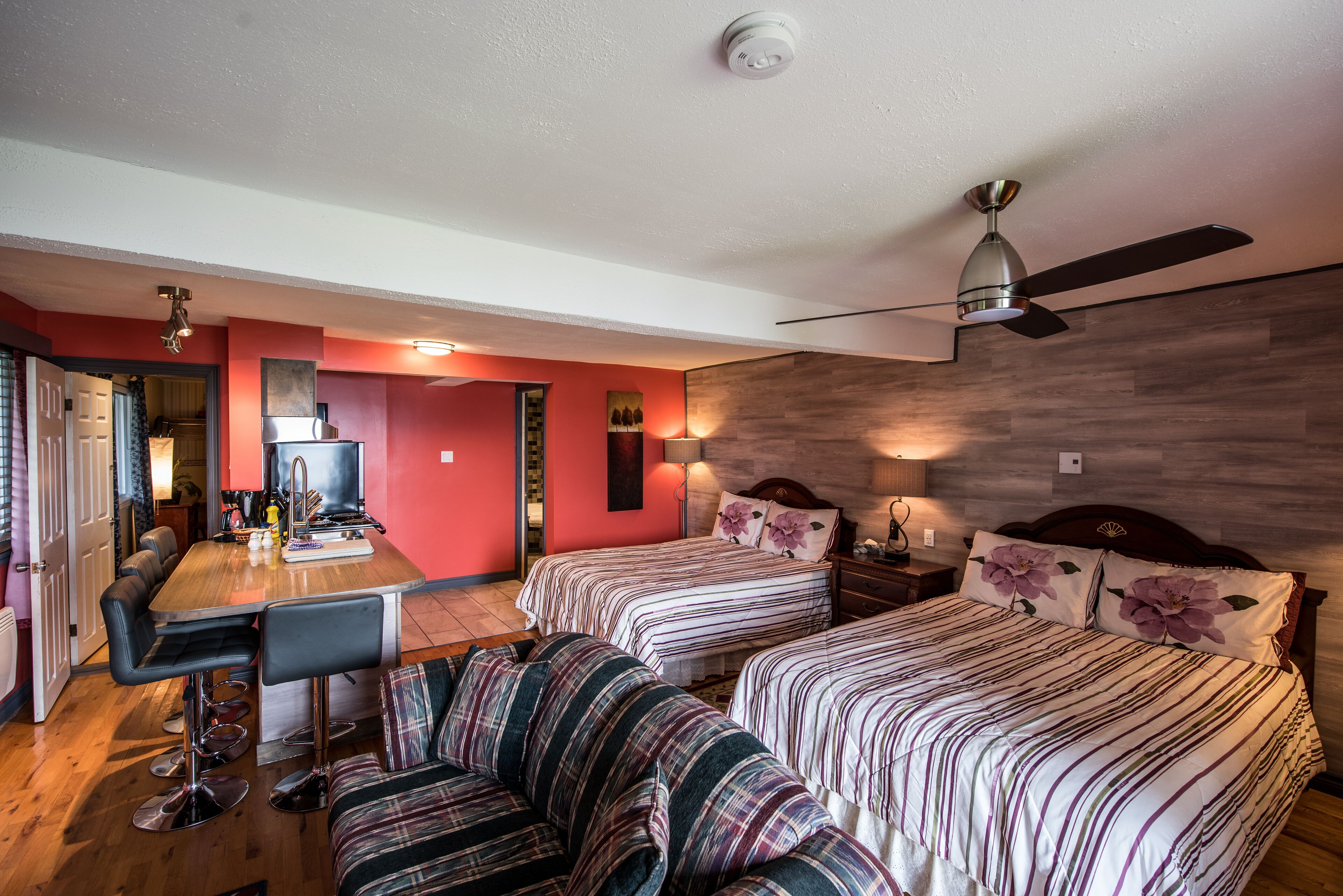 Quarto de hotel moderno com chão de madeira, duas camas de solteiro com almofadas florais, um sofá, um espaço de refeições com balcão e um ventilador de teto.